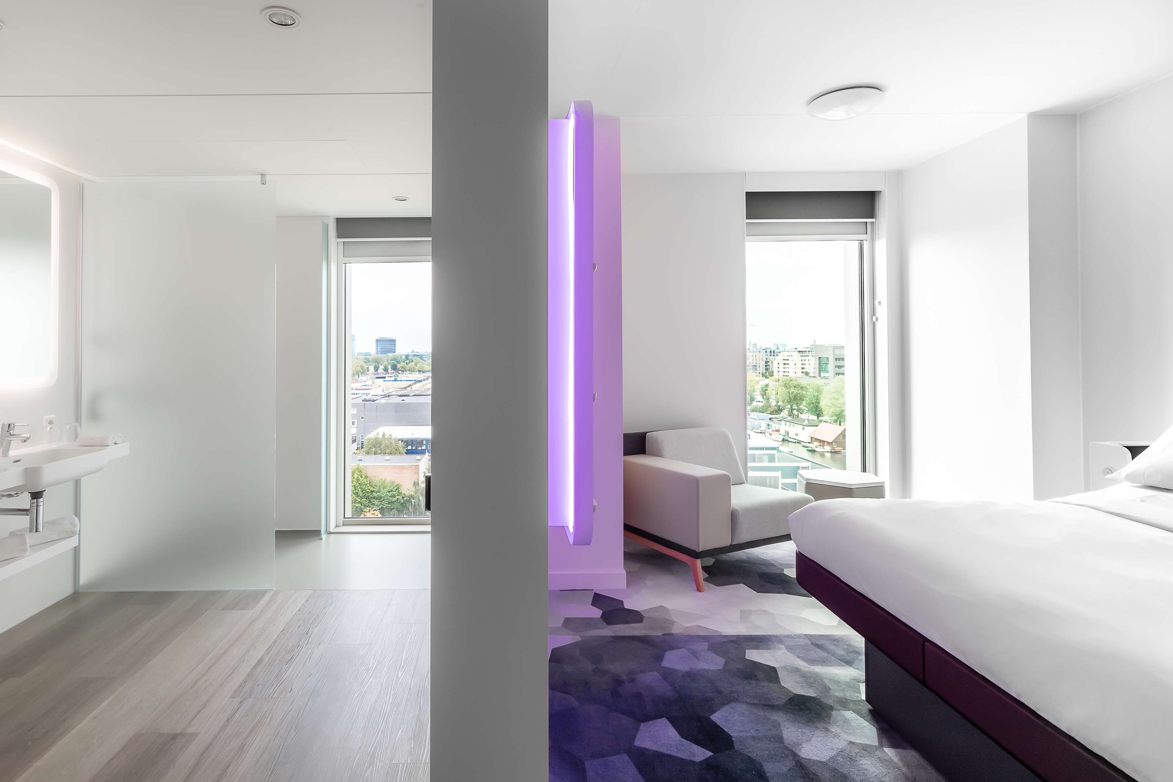 Quarto de hotel moderno com uma cama grande, um espaço de estar com uma poltrona, vista da cidade através de amplas janelas e um pilar iluminado em roxo.