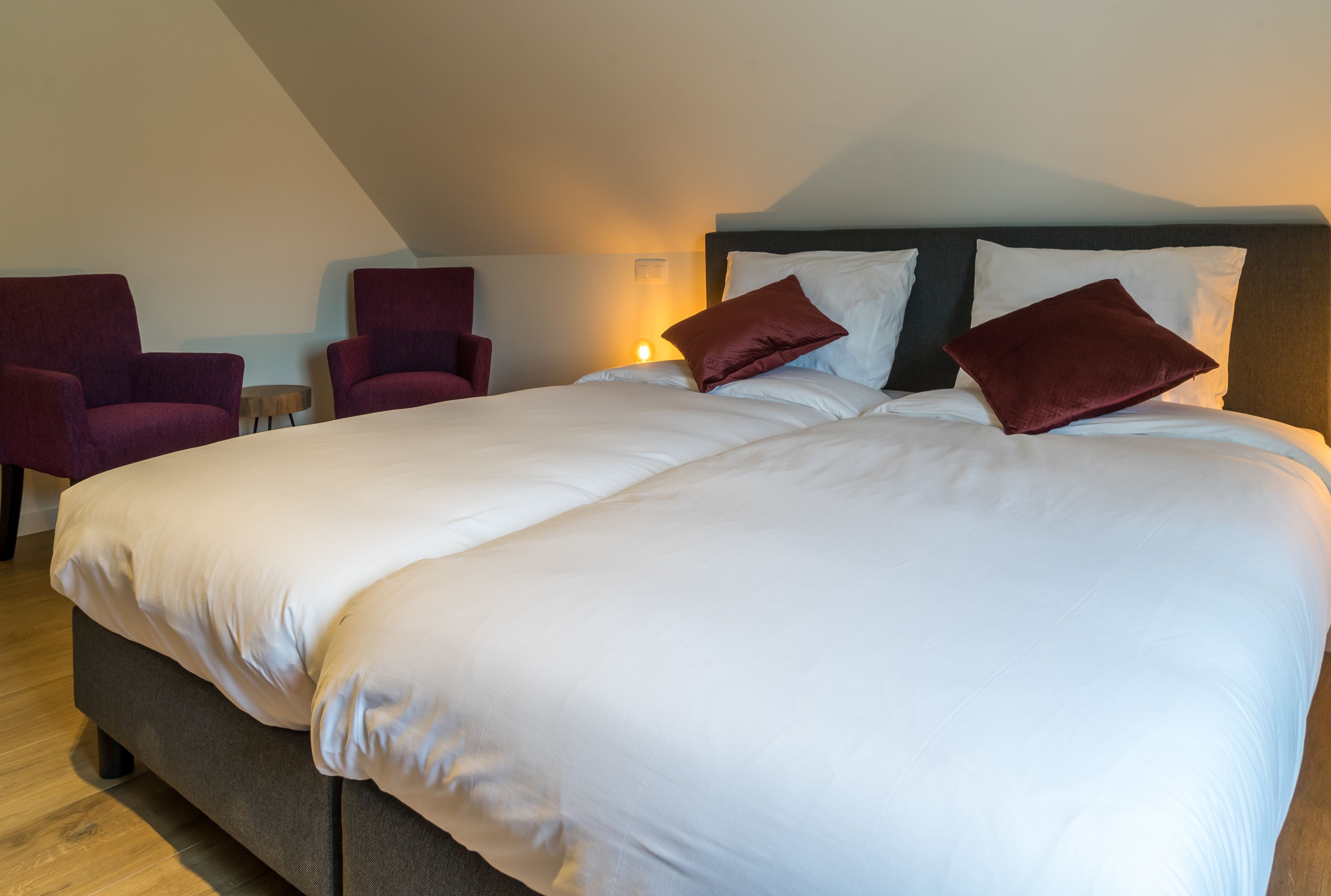Uma cama grande com roupa de cama branca e duas almofadas vermelhas, uma mesa redonda pequena e duas poltronas roxas em um quarto com teto inclinado e piso de madeira.