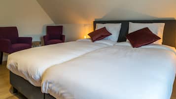 Uma cama grande com roupa de cama branca e duas almofadas vermelhas, uma mesa redonda pequena e duas poltronas roxas em um quarto com teto inclinado e piso de madeira.