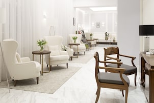 Uma sala de estar moderna com um sofá branco, uma cadeira de madeira e uma mesa de centro. Há uma pintura na parede e um abajur sobre uma mesinha lateral.
