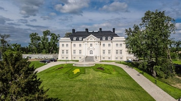 Um edifício grande e imponente, com fachada simétrica, entrada central com degraus e um jardim formal à frente.