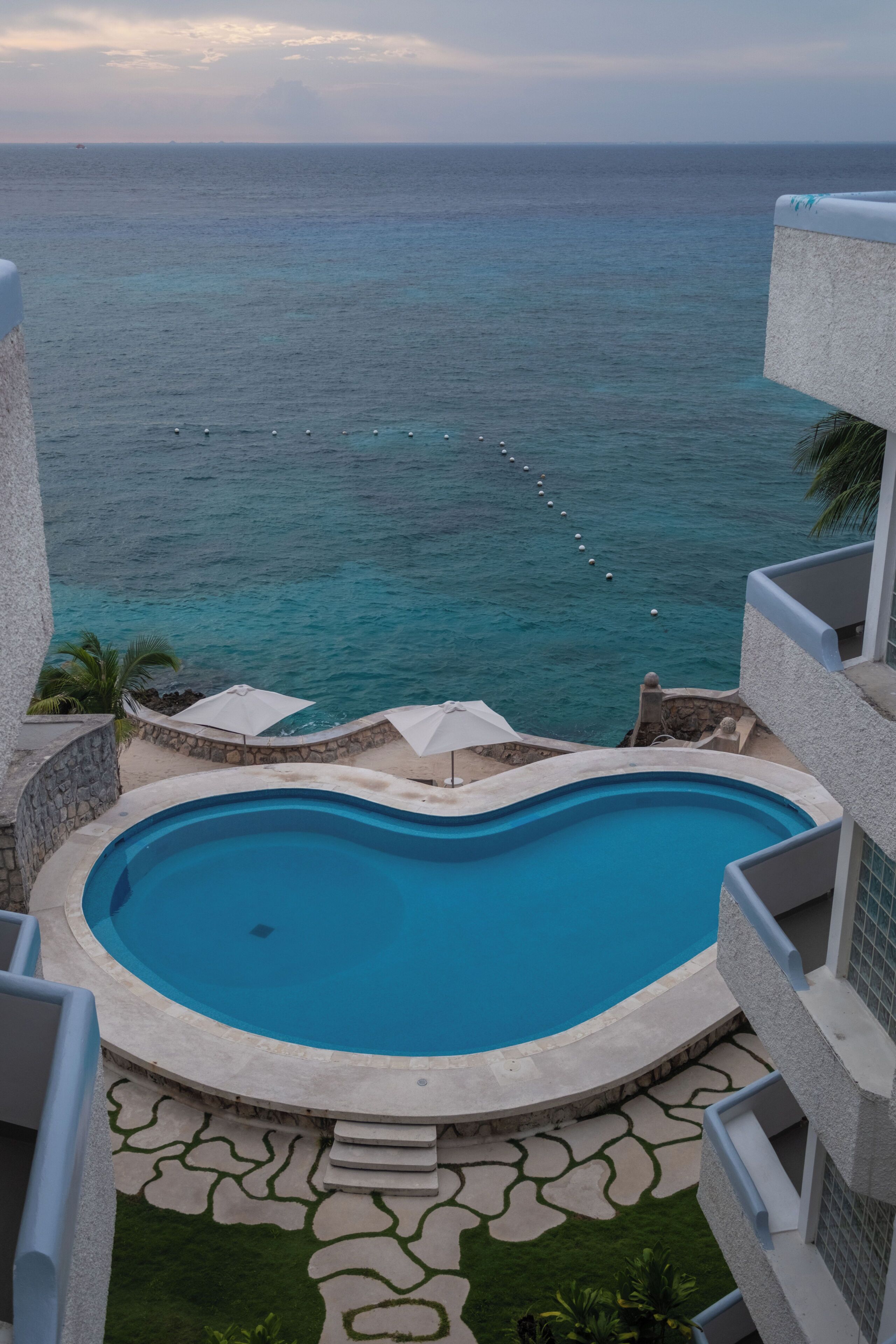 Piscina com borda em formato de coração, rodeada por degraus de pedra e palmeiras, com vista para um corpo de água.