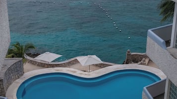 Piscina com borda em formato de coração, rodeada por degraus de pedra e palmeiras, com vista para um corpo de água.