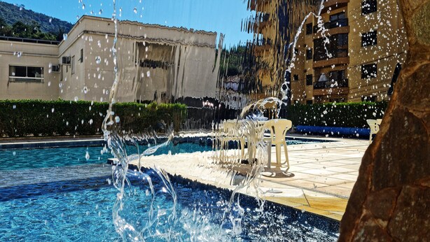 Piscina com jatos de água, estrutura de pedra e um edifício ao fundo.