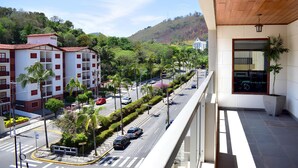 Um hotel de vários andares com varandas, vista da rua com carros e vegetação, e uma paisagem montanhosa ao fundo.
