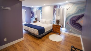 Quarto com cama, piso de madeira, tapete branco, mesa de cabeceira e parede com design abstrato colorido.