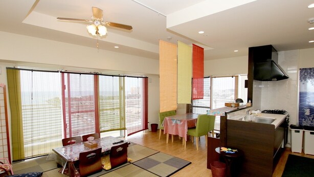 Ampla sala de estar com mesa de jantar, cadeiras, área de cozinha e ventilador de teto.