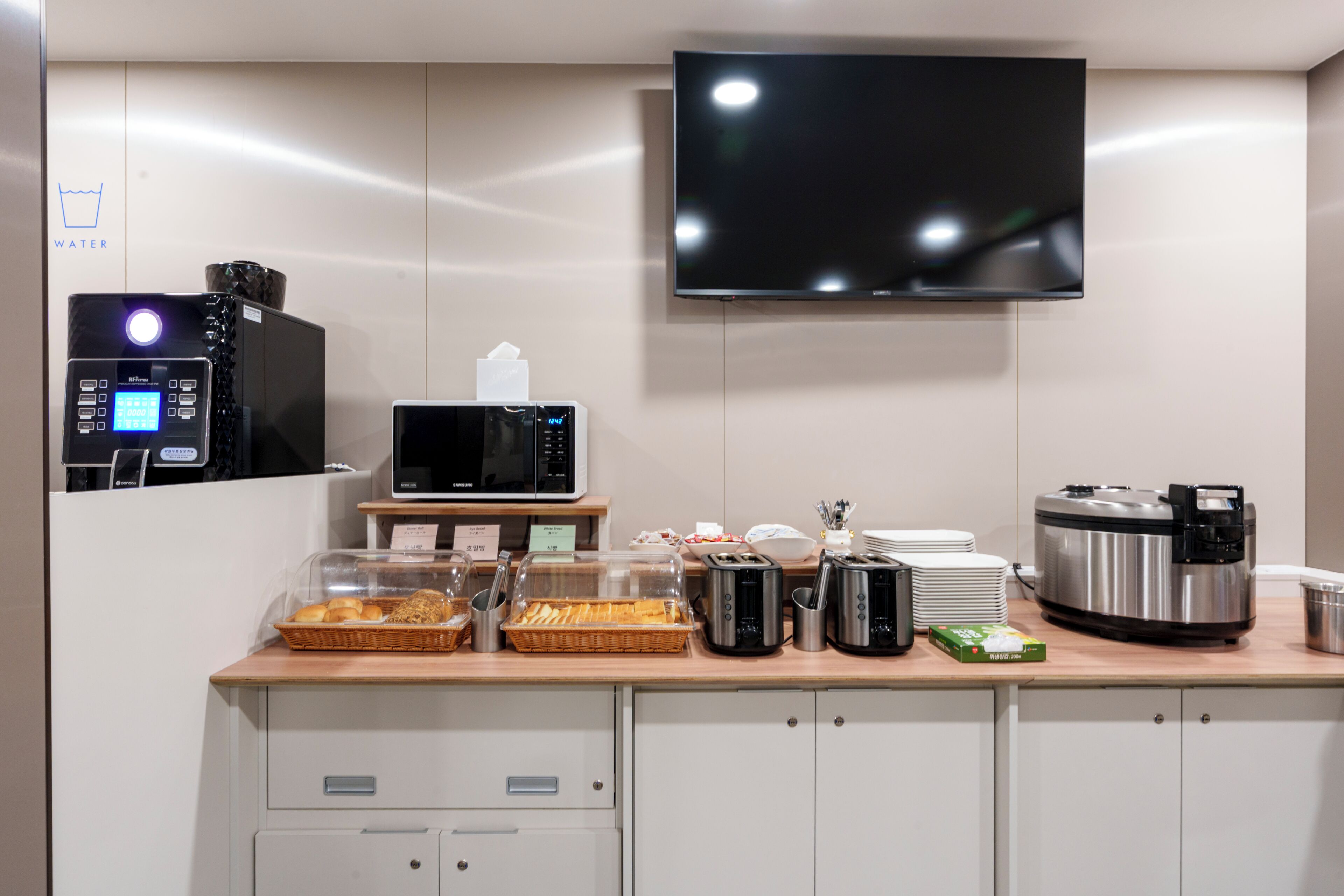 Quarto de hotel com televisão de tela plana, micro-ondas, dispensador de água, mesa de buffet com alimentos e panela elétrica de arroz.