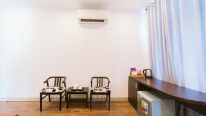 Quarto pequeno e limpo, com piso de madeira, duas cadeiras, uma mesa, um frigorífico pequeno e um aparelho de ar condicionado branco.