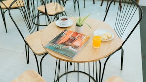Uma mesa de madeira redonda com uma revista, uma chávena de café, um copo de sumo de laranja e uma pequena planta em vaso.