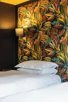 Quarto de hotel com cama, roupa de cama branca e parede com padrão de folhagem tropical.