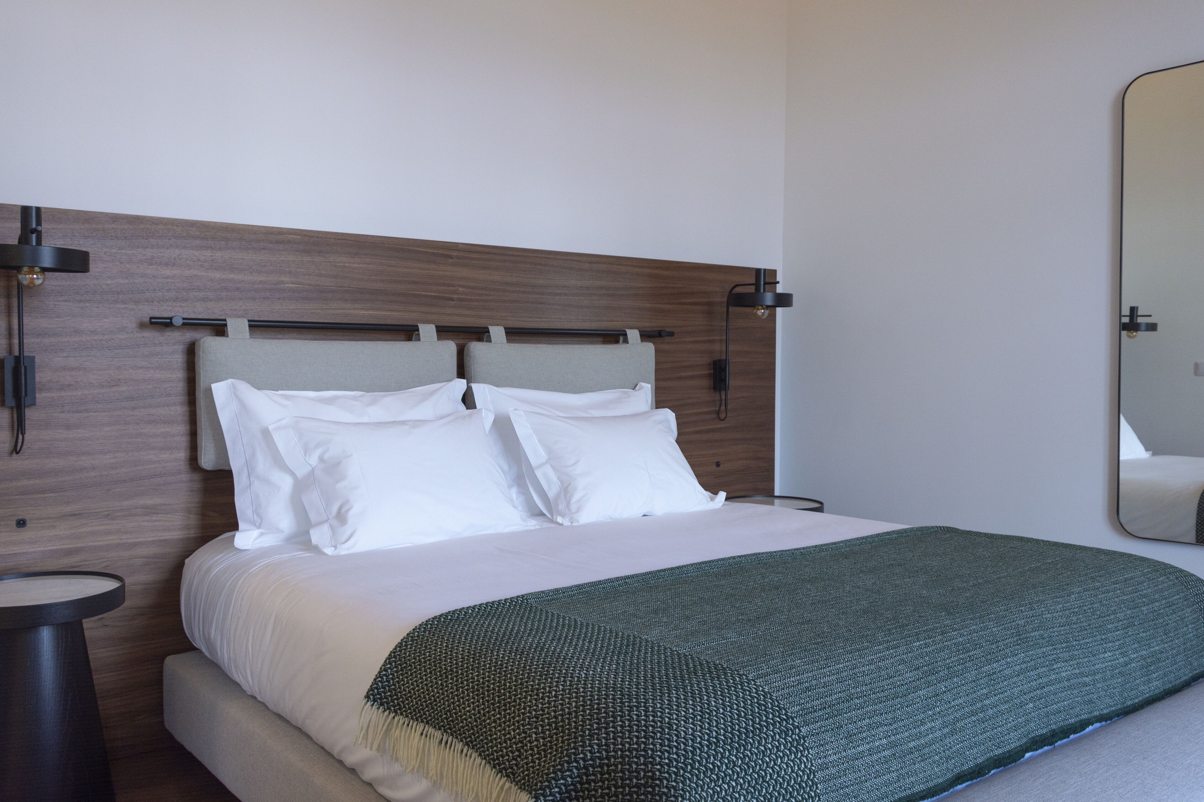 Cama bem arrumada com lençóis brancos e manta verde, cabeceira de madeira e abajures laterais.