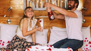 Um casal sentado na cama, com pétalas de rosa espalhadas, segurando uma garrafa e um copo.