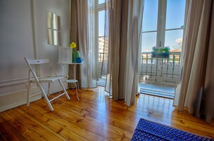 Quarto com piso de madeira, uma cadeira branca, uma mesinha com um bule azul e vista para uma varanda com plantas.