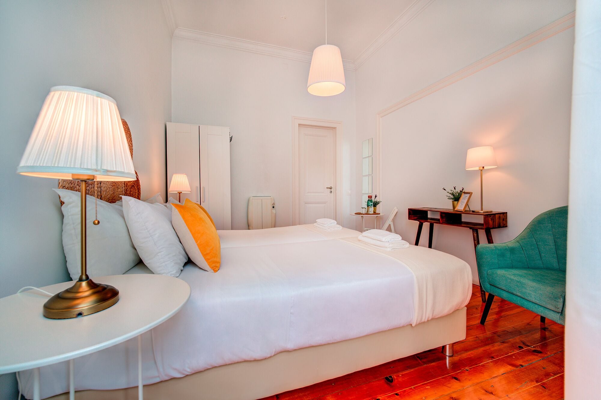 Quarto de hotel com uma cama grande, duas luminárias de cabeceira, uma mesa pequena com uma luminária, uma poltrona verde, piso de madeira e uma porta.