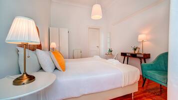 Quarto de hotel com uma cama grande, duas luminárias de cabeceira, uma mesa pequena com uma luminária, uma poltrona verde, piso de madeira e uma porta.
