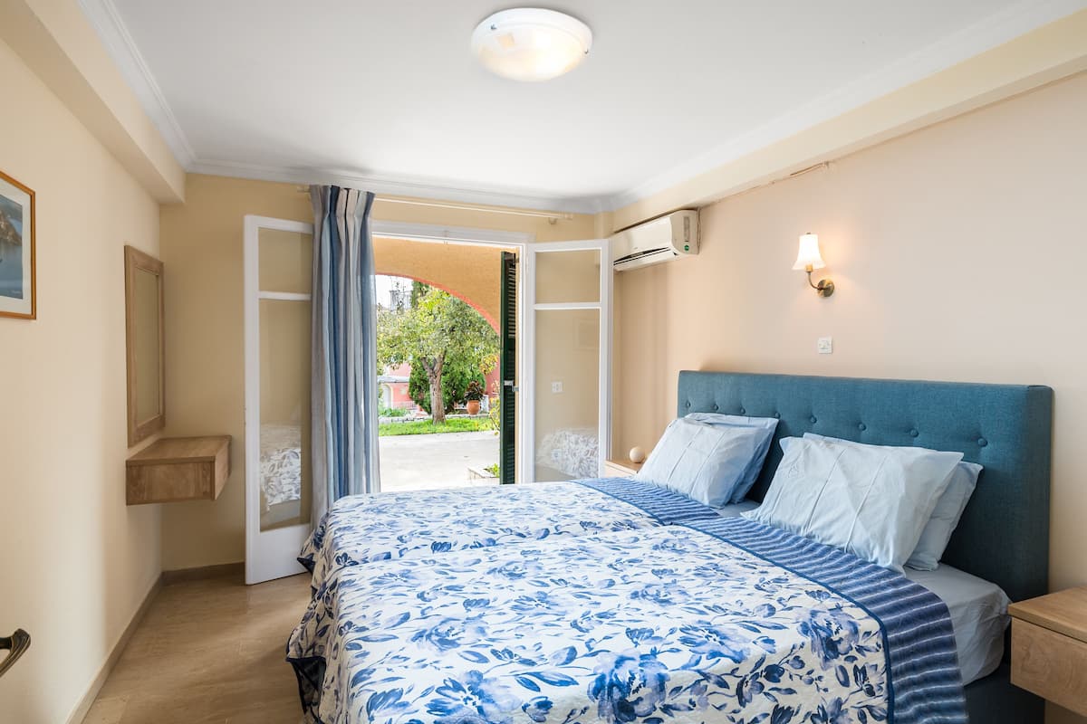 Quarto com cama, mesa de cabeceira, luminária na parede e porta aberta para uma varanda.