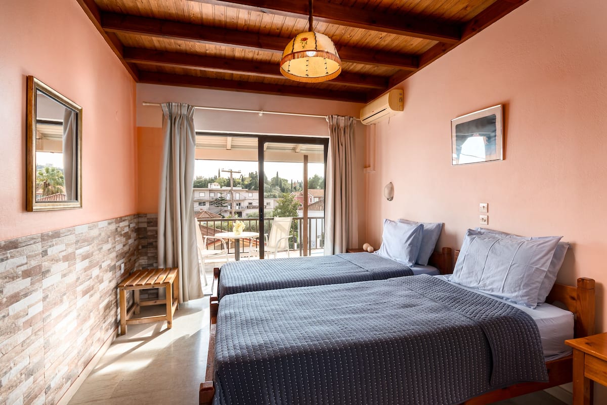 Quarto com duas camas, um banco de madeira, uma varanda com mesa e cadeiras, e vista para prédios.