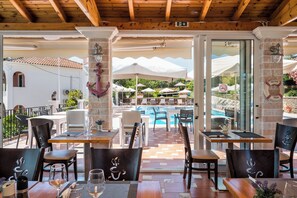 Uma área de refeições ao ar livre com mesas e cadeiras, piscina e guarda-sóis ao fundo.