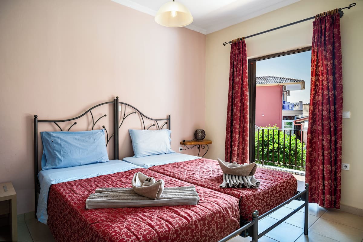 Uma cama de casal com roupa de cama vermelha e branca, uma mesinha de cabeceira com uma luminária e vista para uma varanda com cortinas vermelhas.