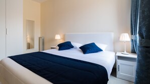 Quarto de hotel com uma cama grande, duas mesas de cabeceira, um criado-mudo, um espelho e uma janela com cortinas.