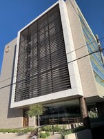 Um edifício moderno com fachada de vidro extensa, um logotipo em destaque à esquerda e uma área ajardinada com plantas e assentos abaixo.