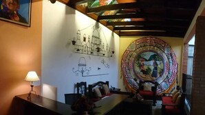 Quarto aconchegante com teto de madeira, cama com travesseiros, escrivaninha com abajur e tapeçaria colorida na parede.