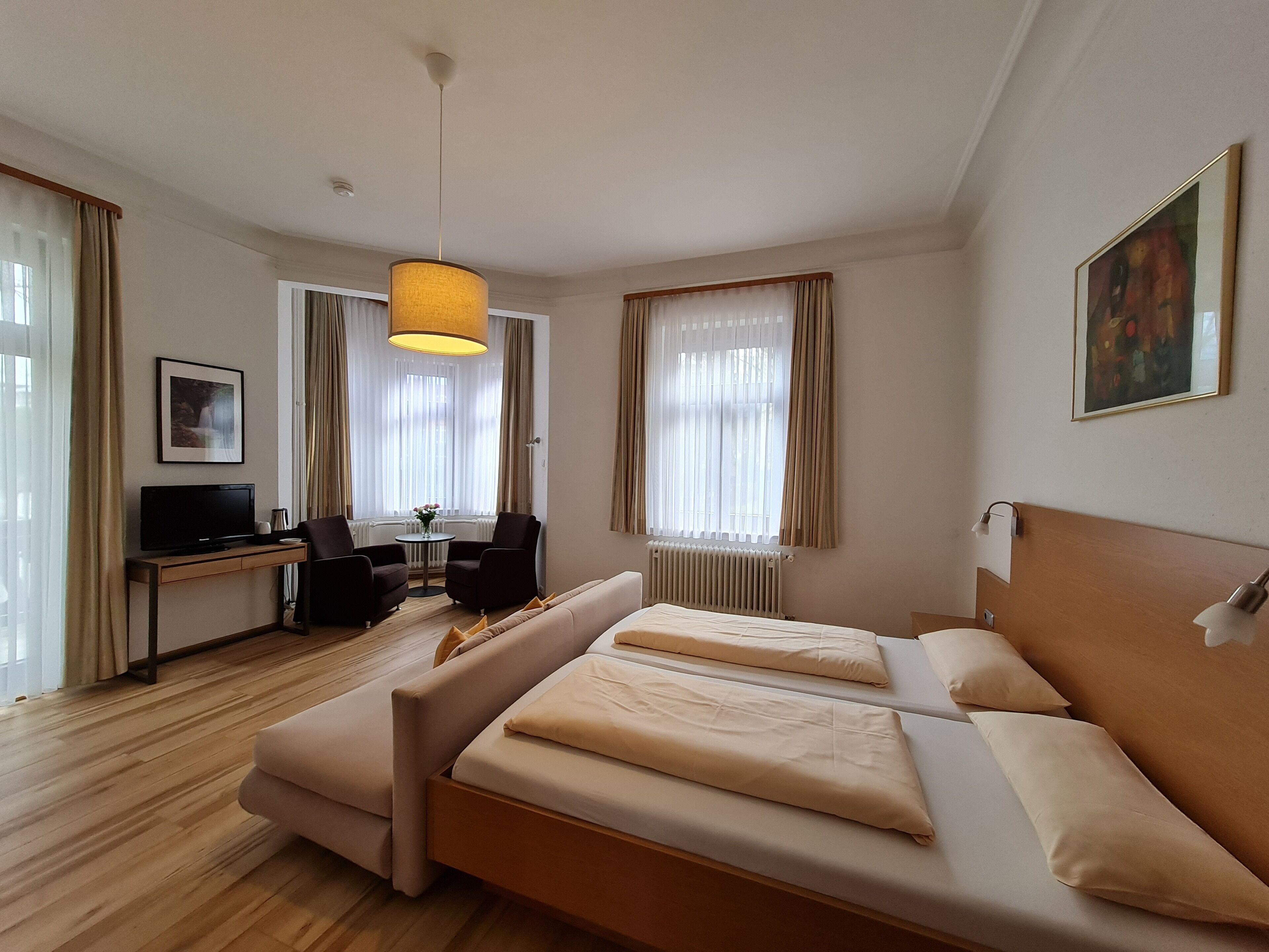 Quarto de hotel com cama, escrivaninha, cadeira, televisão e um quadro na parede.