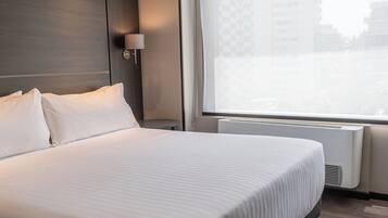 Quarto de hotel moderno com uma cama grande, luminária de cabeceira, janela com vista para edifícios e ar-condicionado fixado na parede.