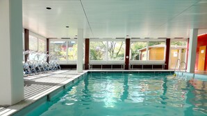 Piscina interna com água cristalina, cercada por janelas e equipamentos de ginástica.