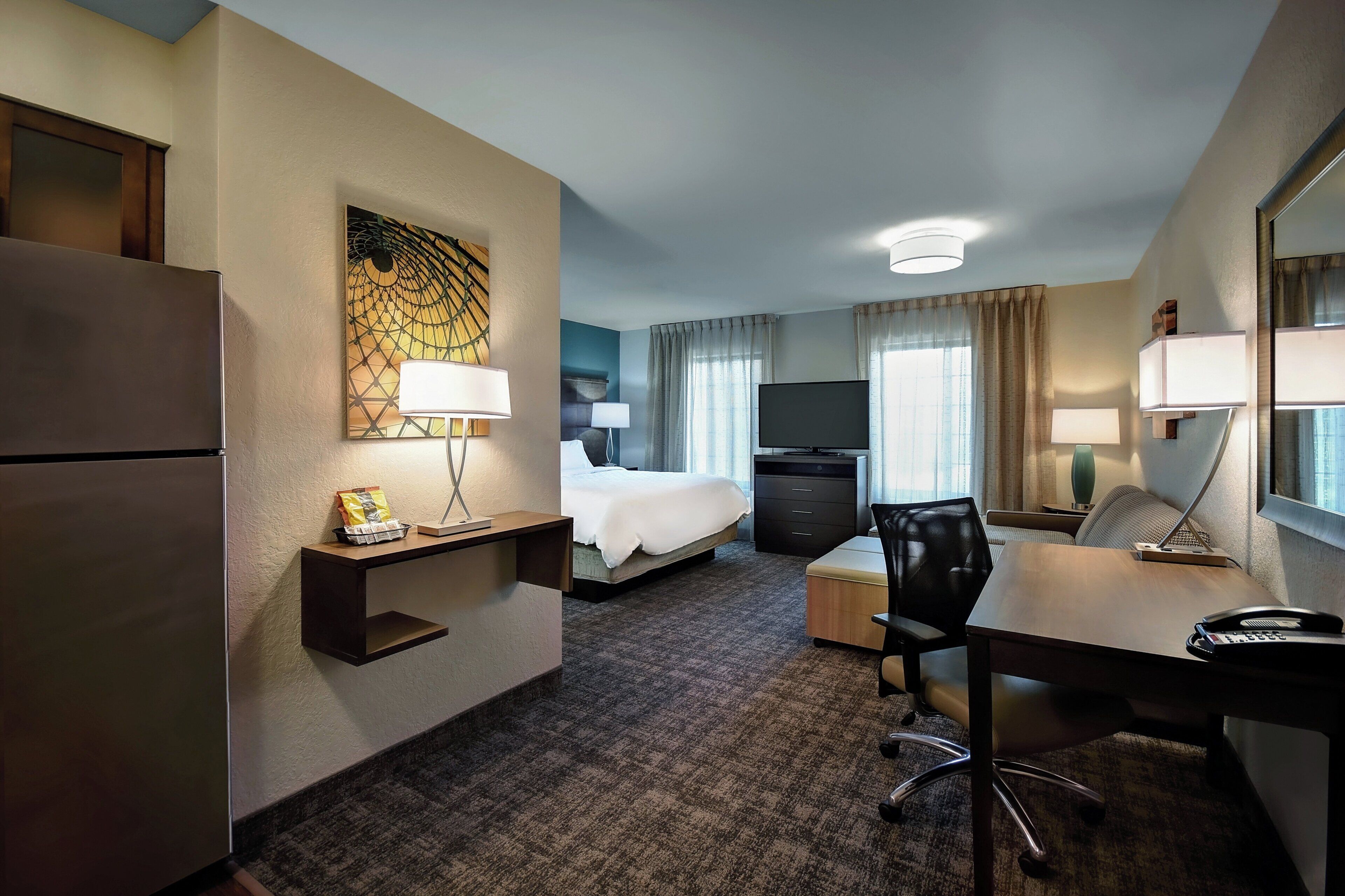 Quarto de hotel com cama, escrivaninha, cadeira, televisão e frigorífico.