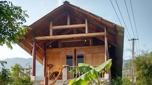 Um edifício rústico de madeira com telhado inclinado, rodeado por vegetação exuberante e um caminho de pedra que leva à entrada.