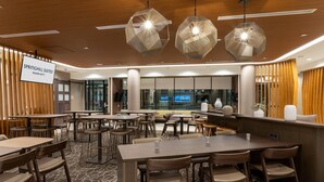 Um hall de hotel moderno com várias mesas e cadeiras para refeições, um balcão de recepção e um grande painel digital.