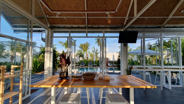 Uma área de refeições coberta ao ar livre, vista para a piscina, com banquetas brancas e mesa de madeira.