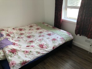 Uma cama de solteiro com roupa de cama floral, uma janela com cortinas e piso de madeira.