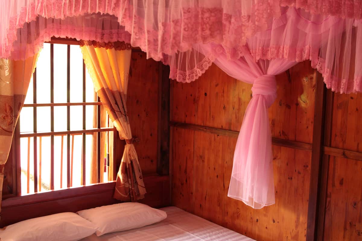 Quarto de madeira com uma cama, janela com grades e cortinas cor-de-rosa.