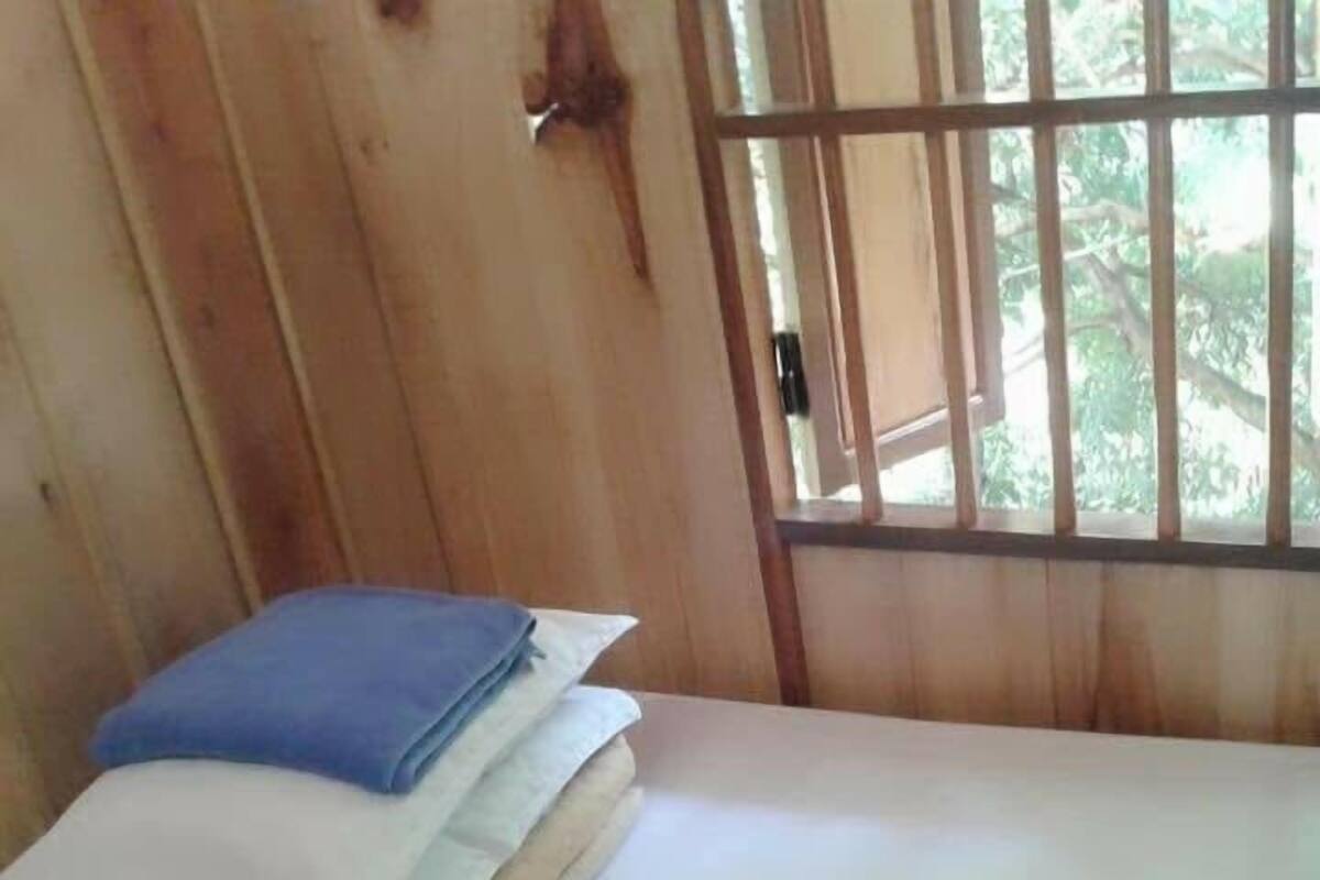 Quarto de madeira com cama, janela com grades e vista para árvores.