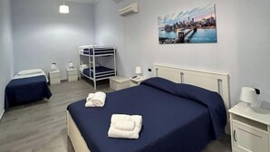 Quarto de hotel com beliche, uma cama de solteiro, pintura de paisagem urbana, luminária no teto e mesa de cabeceira com abajur.