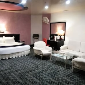 Quarto de hotel com uma cama, duas poltronas brancas, uma mesa de centro de vidro, uma cadeira vermelha, uma escrivaninha preta e um espelho na parede.