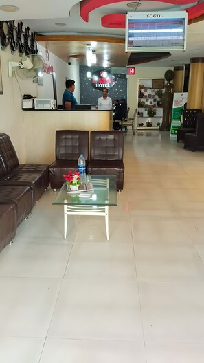 Uma área de recepção de hotel com balcão, dois sofás de couro marrom, uma mesa de centro de vidro com uma garrafa e flores, e uma grande TV de tela plana fixada no teto.
