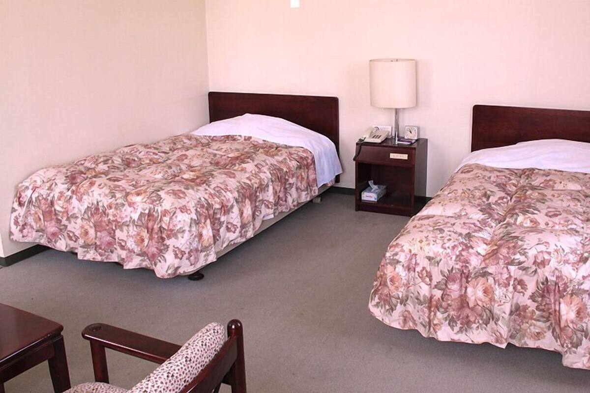 Duas camas de solteiro com roupa de cama floral, uma mesa de cabeceira com abajur e uma cadeira em um quarto de hotel.