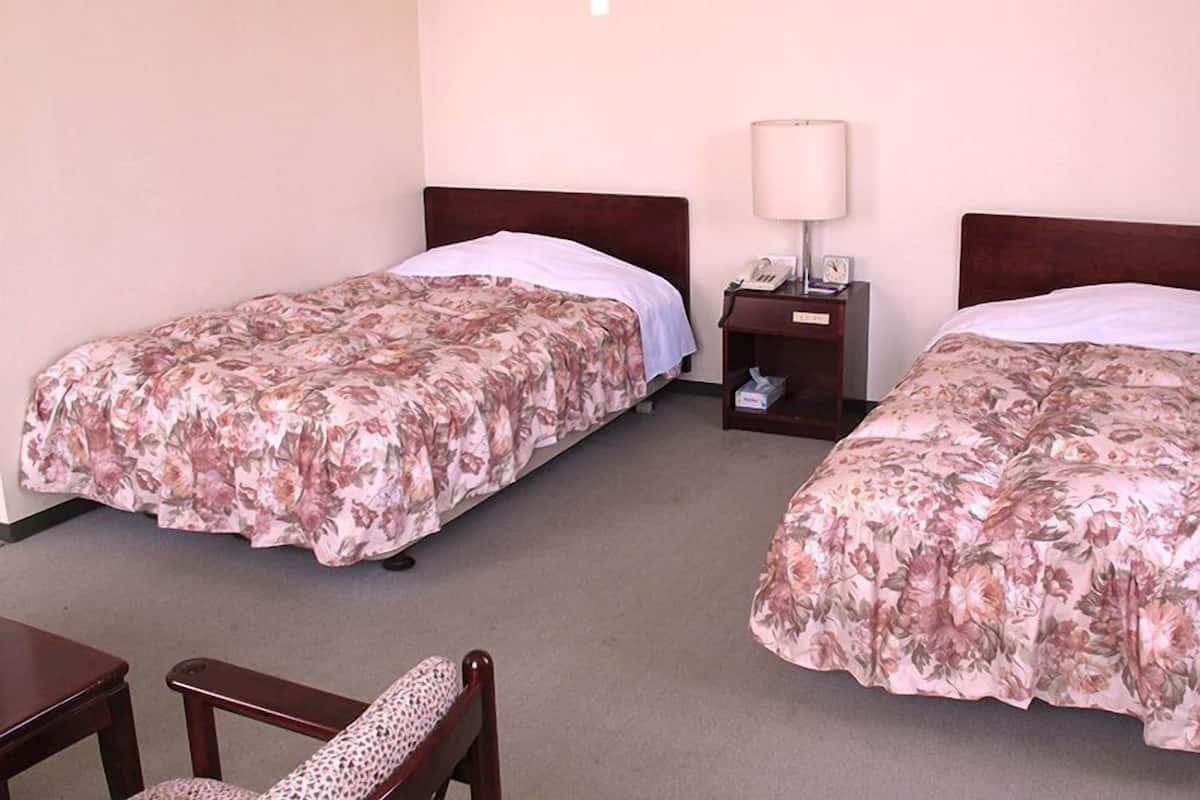 Duas camas de solteiro com roupa de cama floral, uma mesa de cabeceira com abajur e uma cadeira em um quarto de hotel.