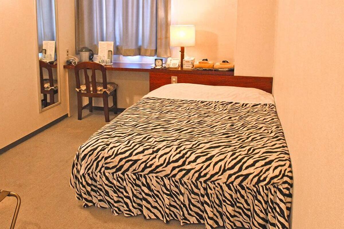 Quarto de hotel com cama, cadeira, escrivaninha, abajur e espelho.