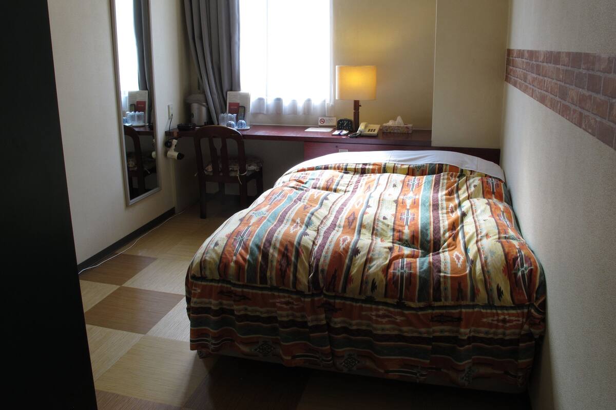 Quarto de hotel com cama, escrivaninha, cadeira, janela com cortinas e luminária.