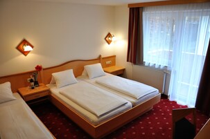 Quarto de hotel com duas camas, cabeceira de madeira, mesa de cabeceira com abajur, janela com cortinas e radiador.