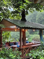 Um gazebo de madeira com telhado de metal corrugado, rodeado por vegetação exuberante e vista para uma encosta arborizada.