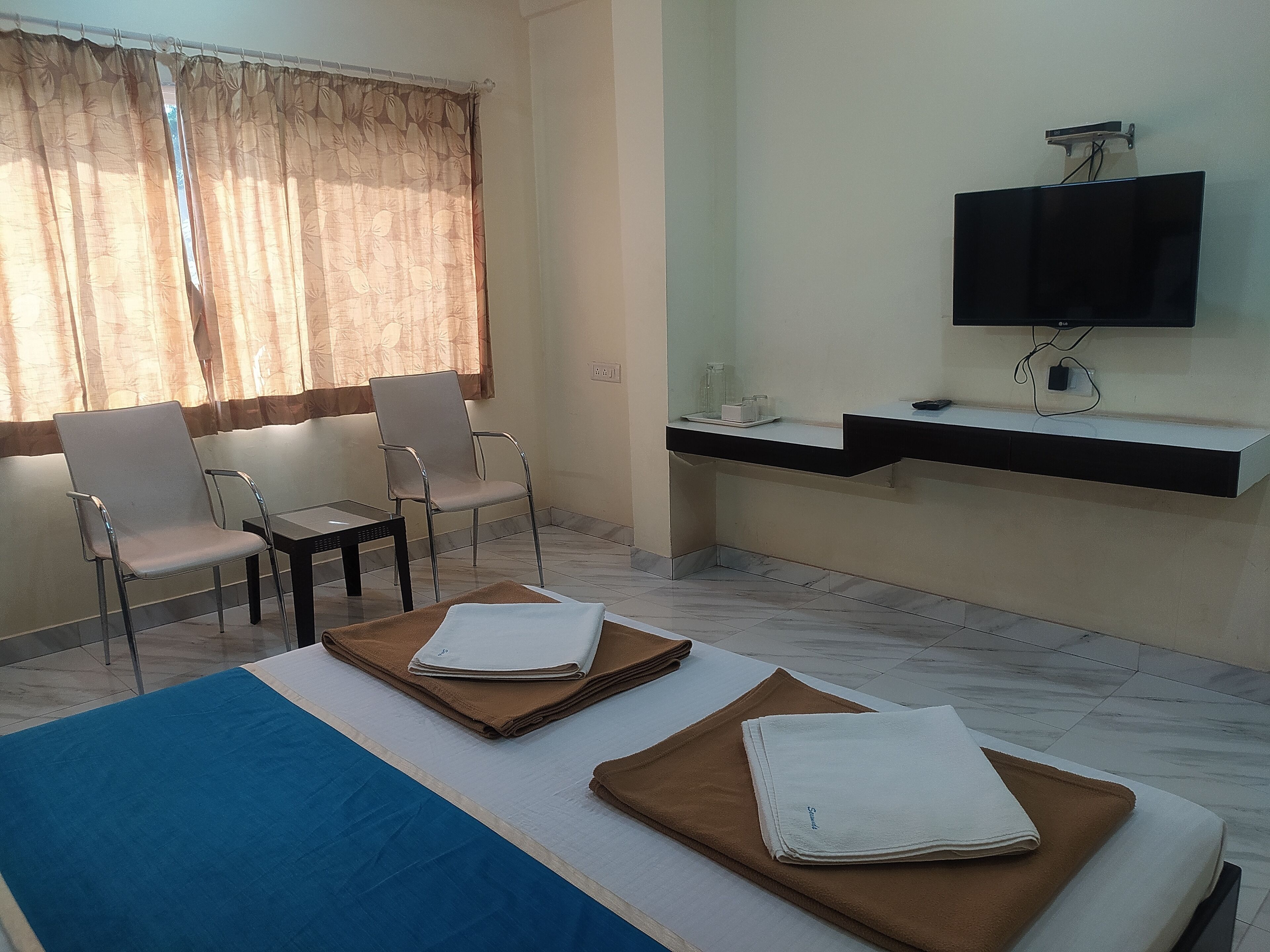 Quarto de hotel com uma cama, duas cadeiras, uma mesa pequena, uma televisão e uma estante com um espelho.