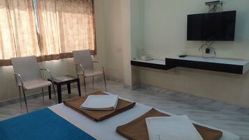 Quarto de hotel com uma cama, duas cadeiras, uma mesa pequena, uma televisão e uma estante com um espelho.