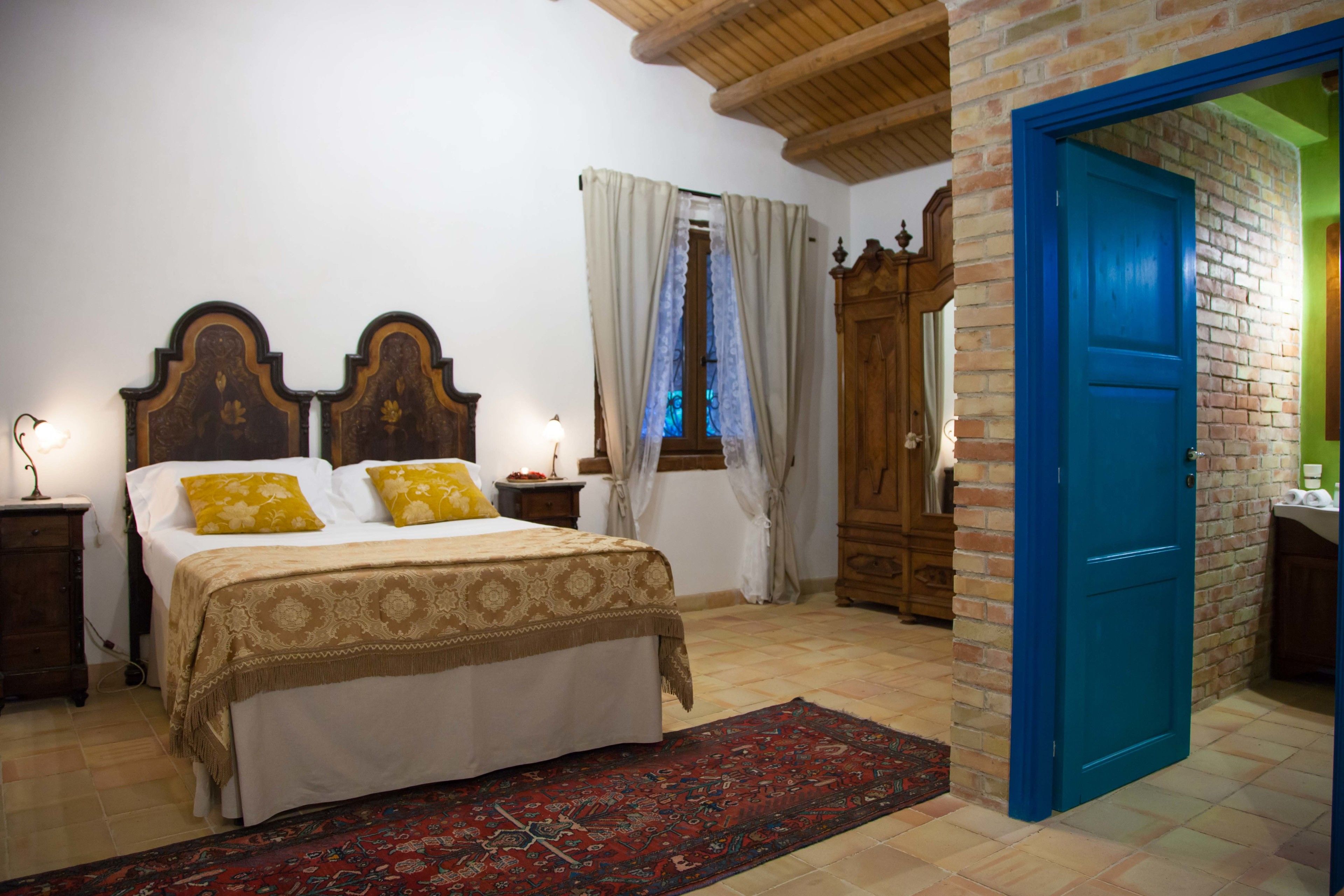 Quarto com cama de casal, cabeceira de madeira, tapete vermelho estampado, porta azul e janela com cortinas.