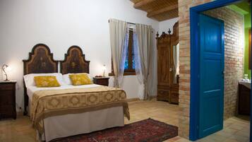 Quarto com cama de casal, cabeceira de madeira, tapete vermelho estampado, porta azul e janela com cortinas.